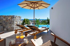 Uma área de estar ao ar livre ensolarada, com cadeiras de lounge e uma mesa, um guarda-sol grande e vista para uma piscina e o horizonte da cidade.