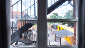 Vista pela janela com uma escadaria de metal que leva para o exterior, um edifício amarelo e uma parede de tijolos.