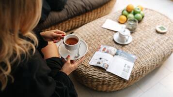 Uma pessoa segurando uma xícara de chá sobre um banquinho de fibra natural, com uma revista, um copo de água e uma tigela de frutas.
