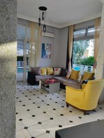 Uma sala de estar moderna com um sofá cinzento, uma poltrona amarela e uma mesa de centro. Há uma janela grande com cortinas, um aparelho de ar condicionado na parede e um quadro sobre o sofá.