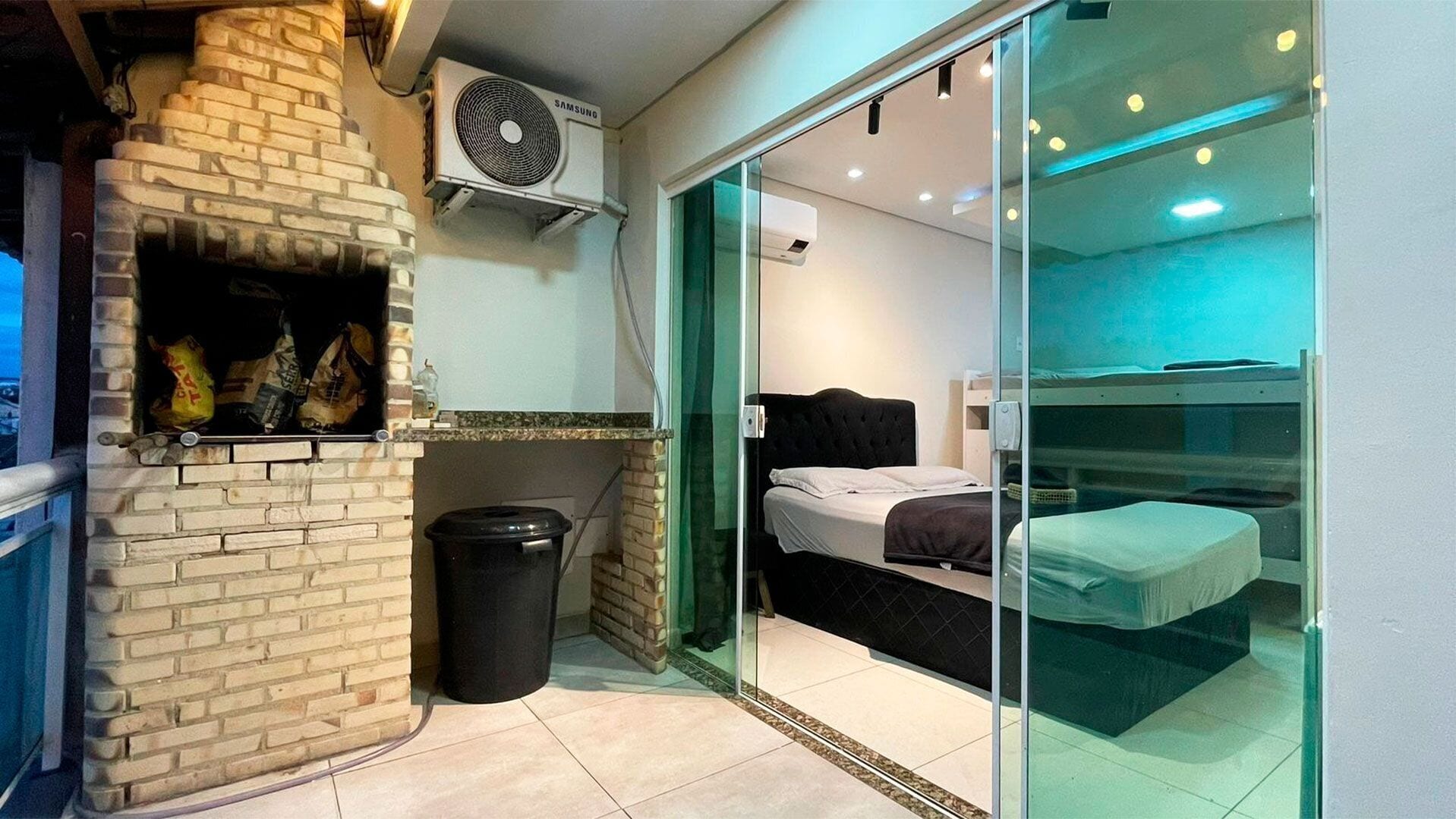 Quarto com lareira de tijolos, uma cama, porta de vidro que dá para uma varanda e ar-condicionado Samsung.