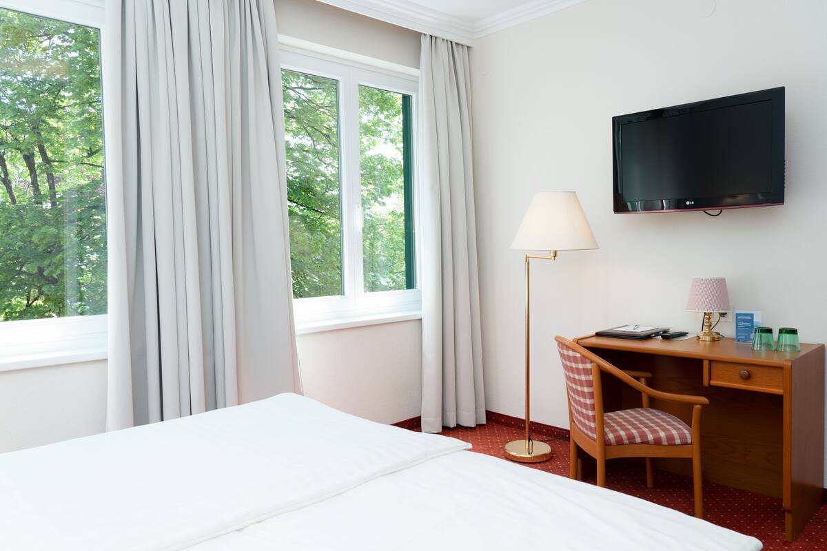 Quarto de hotel com cama, escrivaninha, cadeira, abajur e televisão.