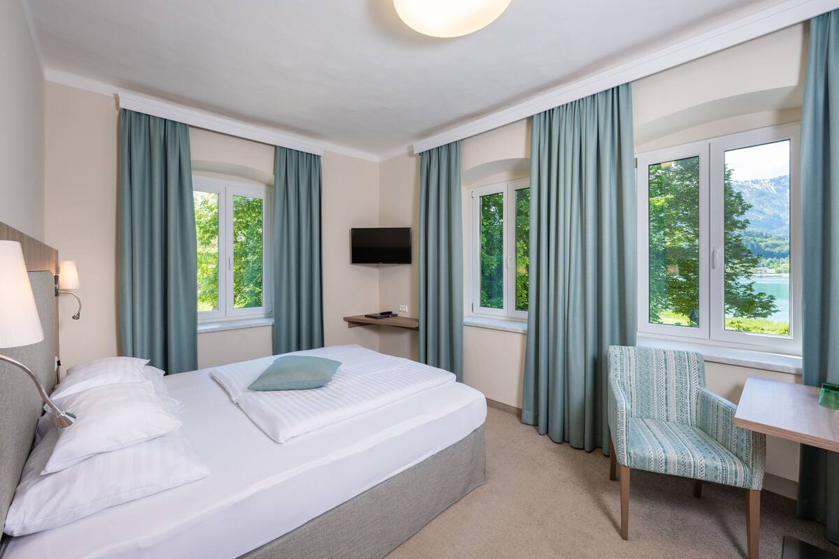 Quarto de hotel com cama, uma cadeira, televisão e janelas com cortinas.