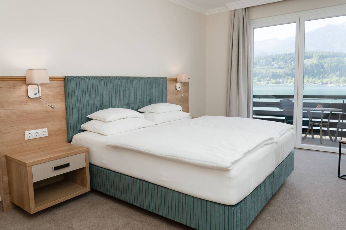 Quarto de hotel com uma cama grande, uma mesa de cabeceira de madeira, duas luminárias na parede e uma varanda com vista para um lago e montanhas.