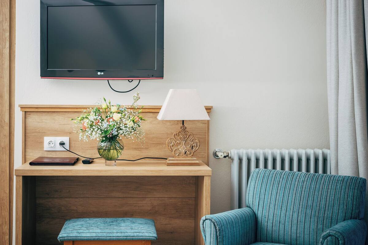 Quarto de hotel com secretária de madeira, TV fixada na parede, luminária, vaso com flores, radiador e poltrona listrada.