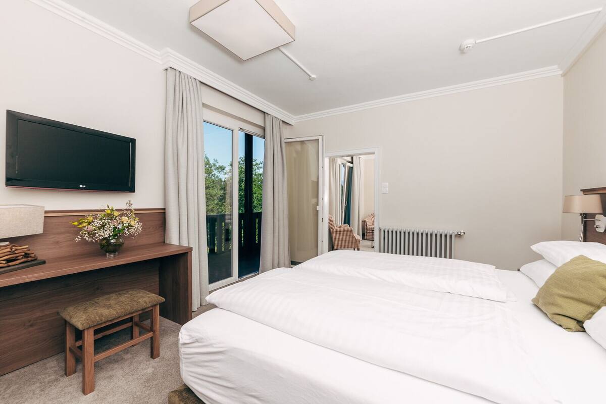 Quarto de hotel com uma cama grande, uma mesa de trabalho com televisão, uma cadeira e vista para o exterior.