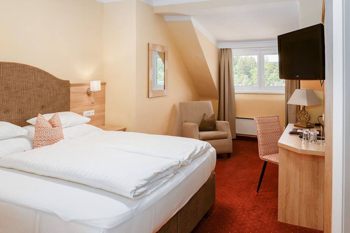 Quarto de hotel com uma cama grande, uma escrivaninha com televisão, uma cadeira e uma janela com cortinas.