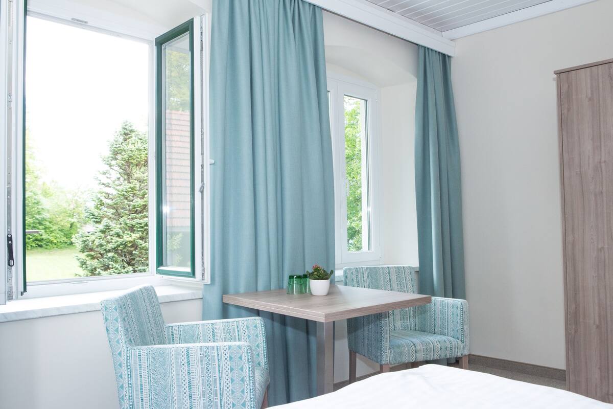 Um quarto com cama, uma mesa, duas cadeiras, janela com cortinas e um guarda-roupa.