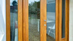 Uma porta de vidro deslizante com moldura de madeira, que dá para uma área externa com uma cadeira de plástico branca e vista para o jardim.