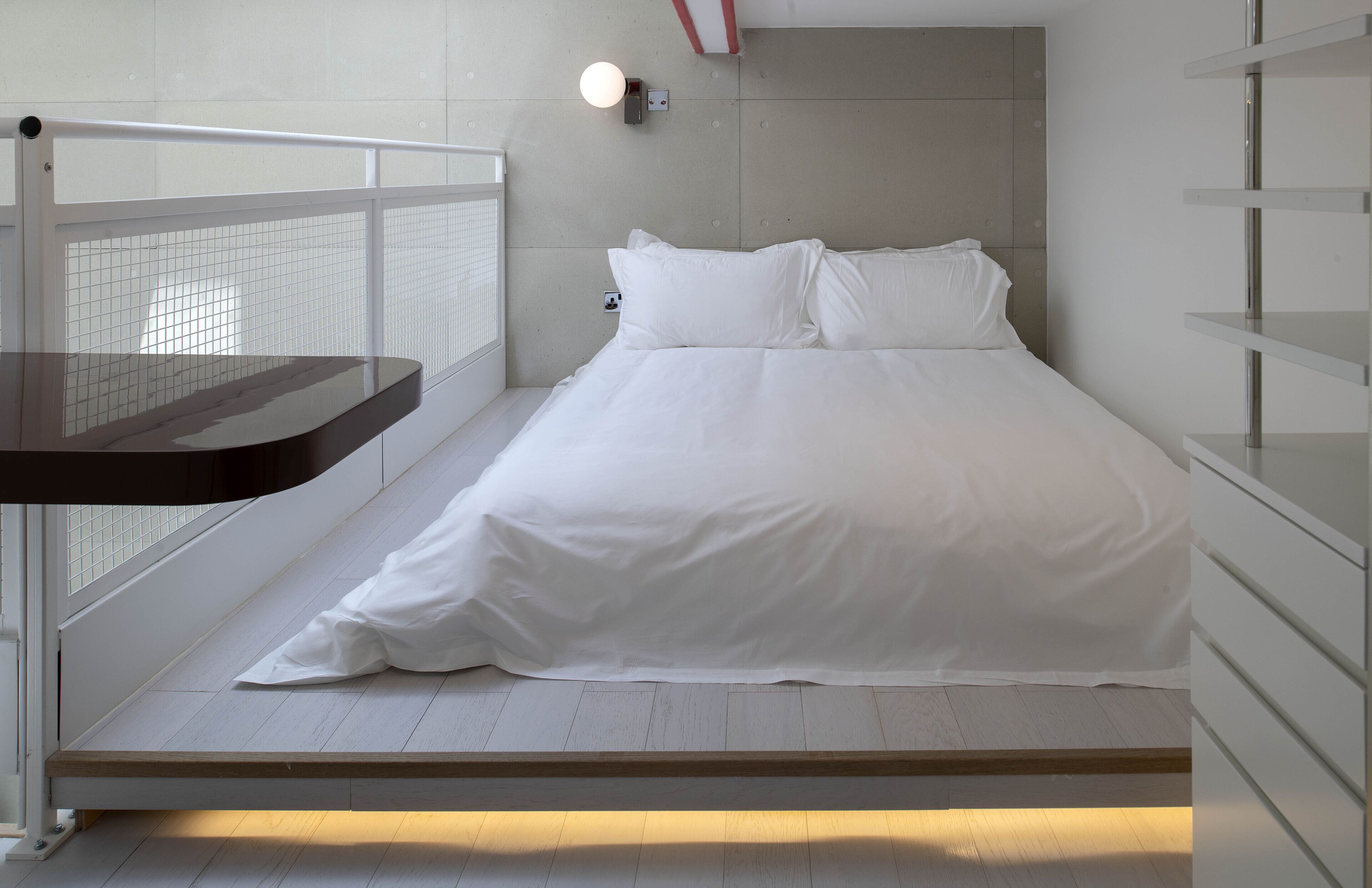 Uma cama em quarto com teto branco, luminária fixada na parede e grade branca.