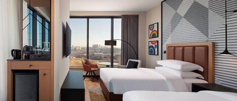 Quarto de hotel com duas camas, uma escrivaninha, uma cadeira, televisão e uma grande janela com vista para a cidade.