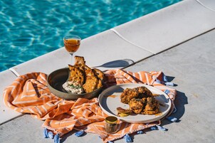 Uma mesa de jantar com um prato de frango frito e uma salada, montada numa praia com uma piscina ao fundo.