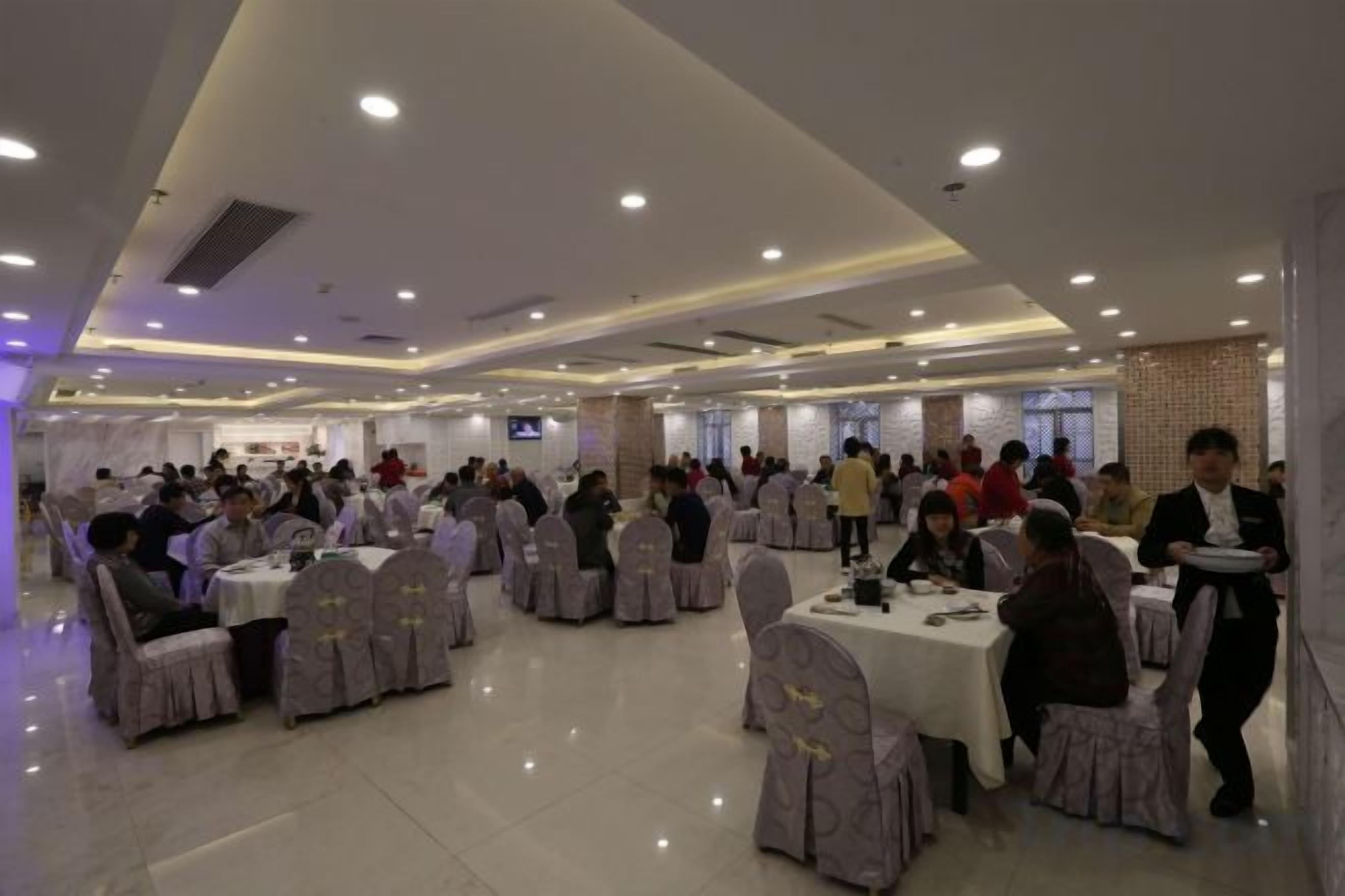 Uma sala de banquetes com mesas e cadeiras redondas, um garçom de uniforme e um grupo de pessoas sentadas.