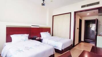 Quarto de hotel com duas camas, uma escrivaninha de madeira, cabeceira vermelha e porta para um banheiro.