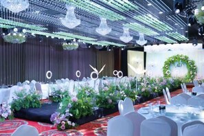 Salão de festas com mesas preparadas para um evento formal, palco com arranjos florais e teto decorativo.