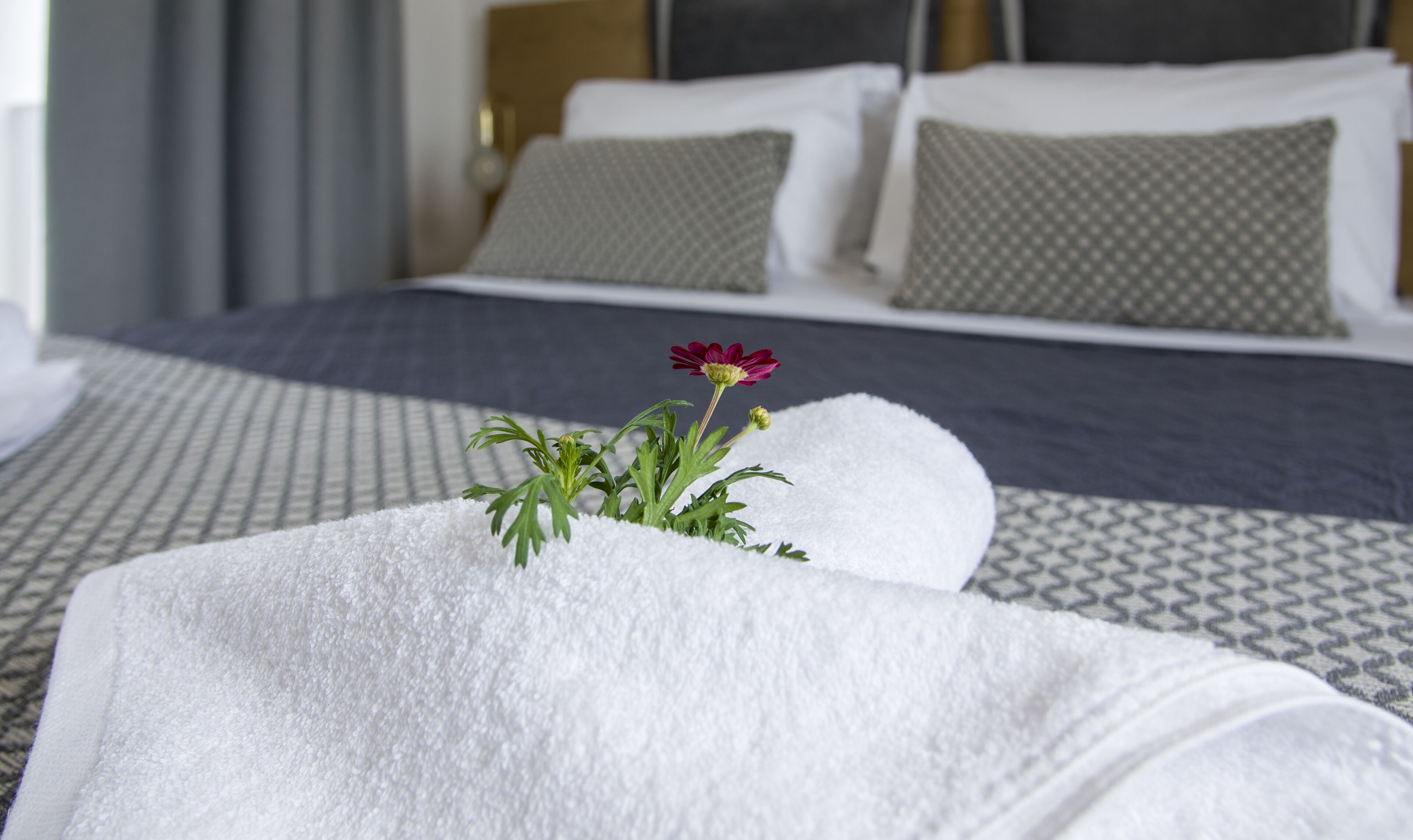 Cama arrumada com colcha estampada, travesseiros e uma única flor sobre uma toalha branca.