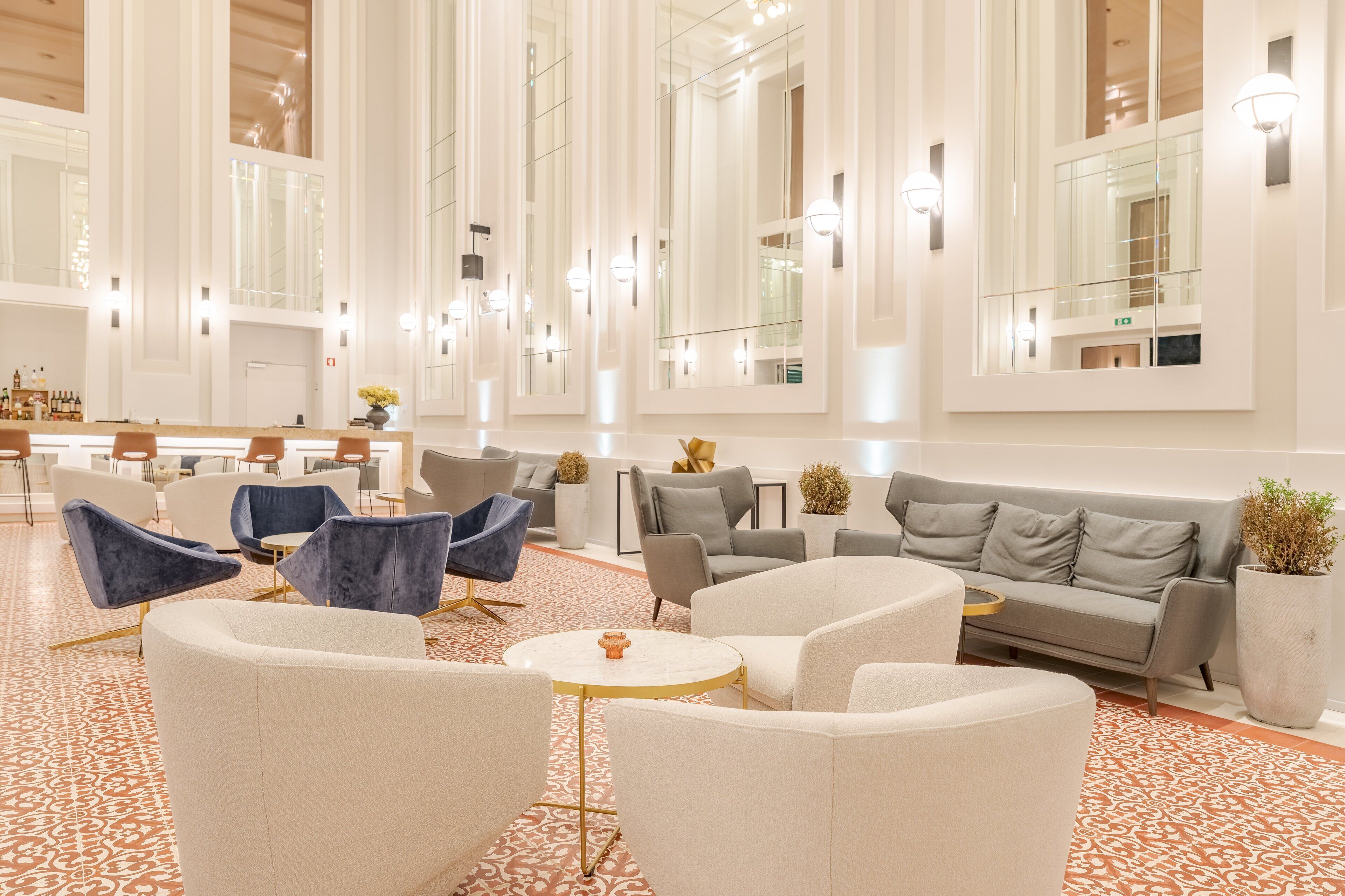 Um hall amplo com diversas opções de assento, incluindo sofás e cadeiras, uma área de bar ao fundo e grandes janelas que permitem a entrada de luz natural.