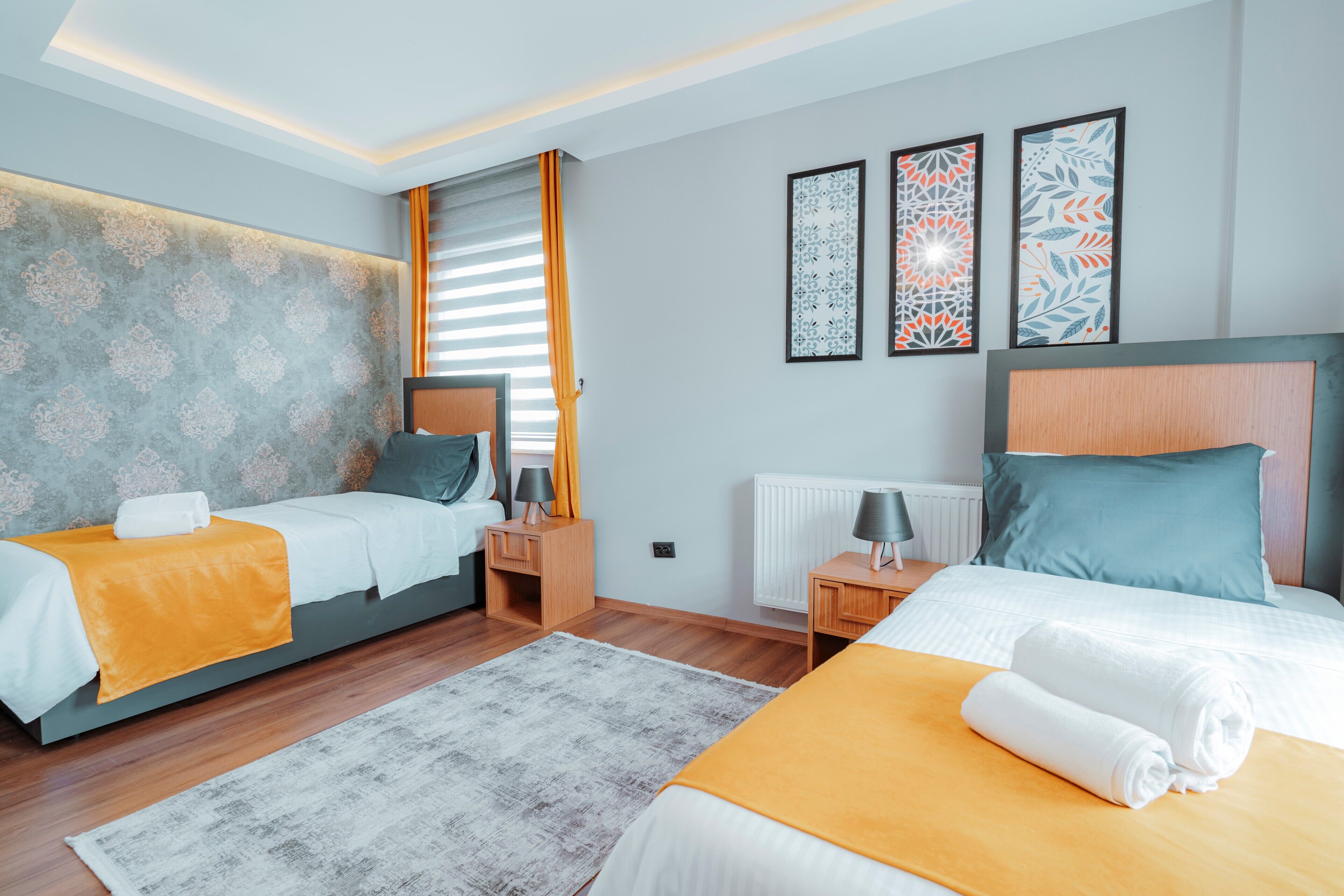 Quarto de hotel com duas camas, uma janela grande com persianas, um radiador e três obras de arte emolduradas na parede.
