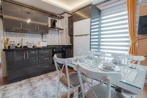 Cozinha moderna com armários escuros, bancada branca e área de refeições para quatro pessoas, com cadeiras brancas e mesa de vidro.