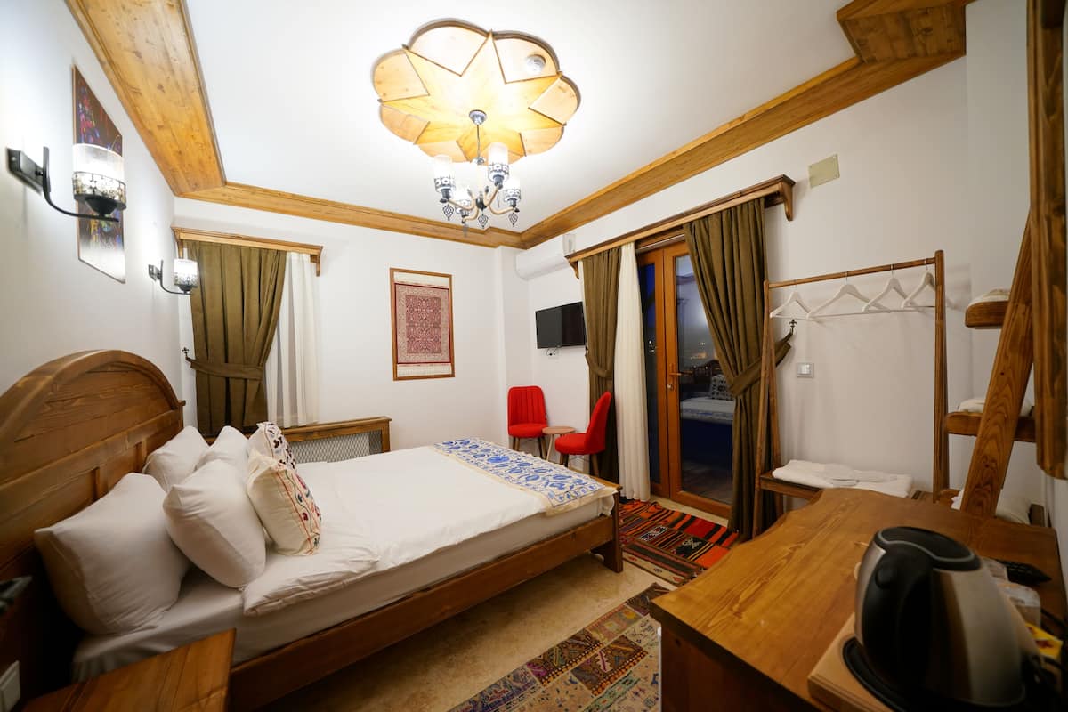 Quarto com cama de madeira, escrivaninha com chaleira, cadeira vermelha e janela grande com cortinas.