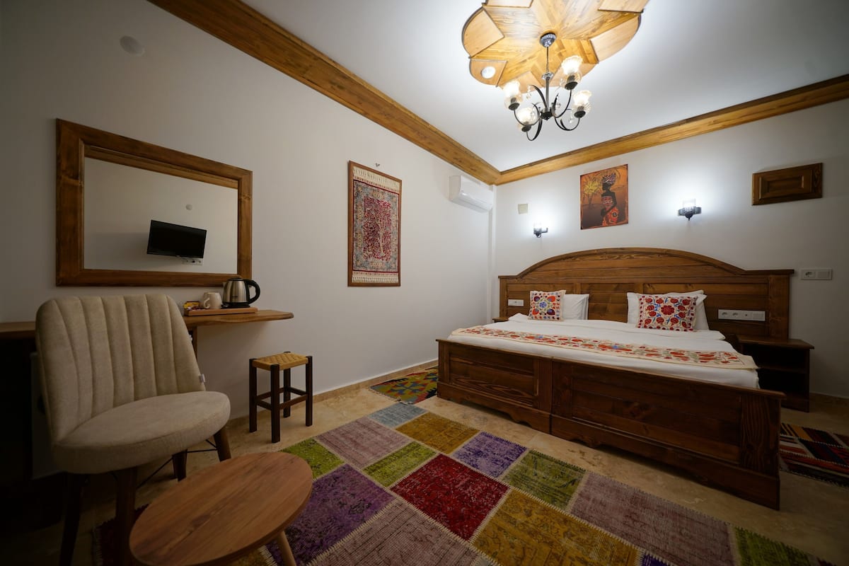 Quarto com cama de madeira, uma cadeira, uma mesinha, um tapete colorido, luminária no teto e obras de arte nas paredes.