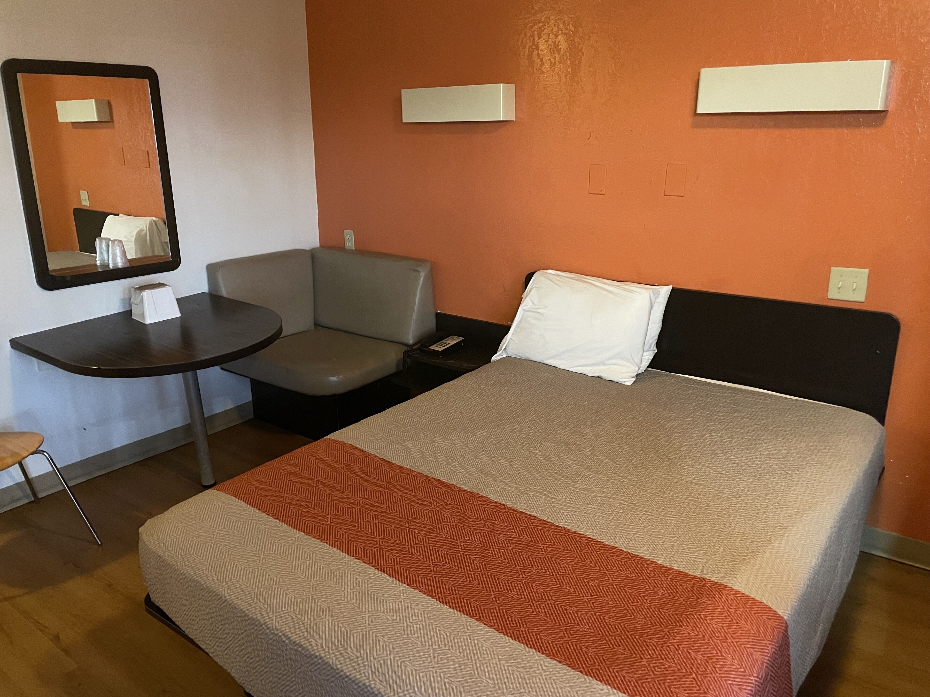 Quarto de hotel com cama, uma mesinha, uma cadeira e um espelho.