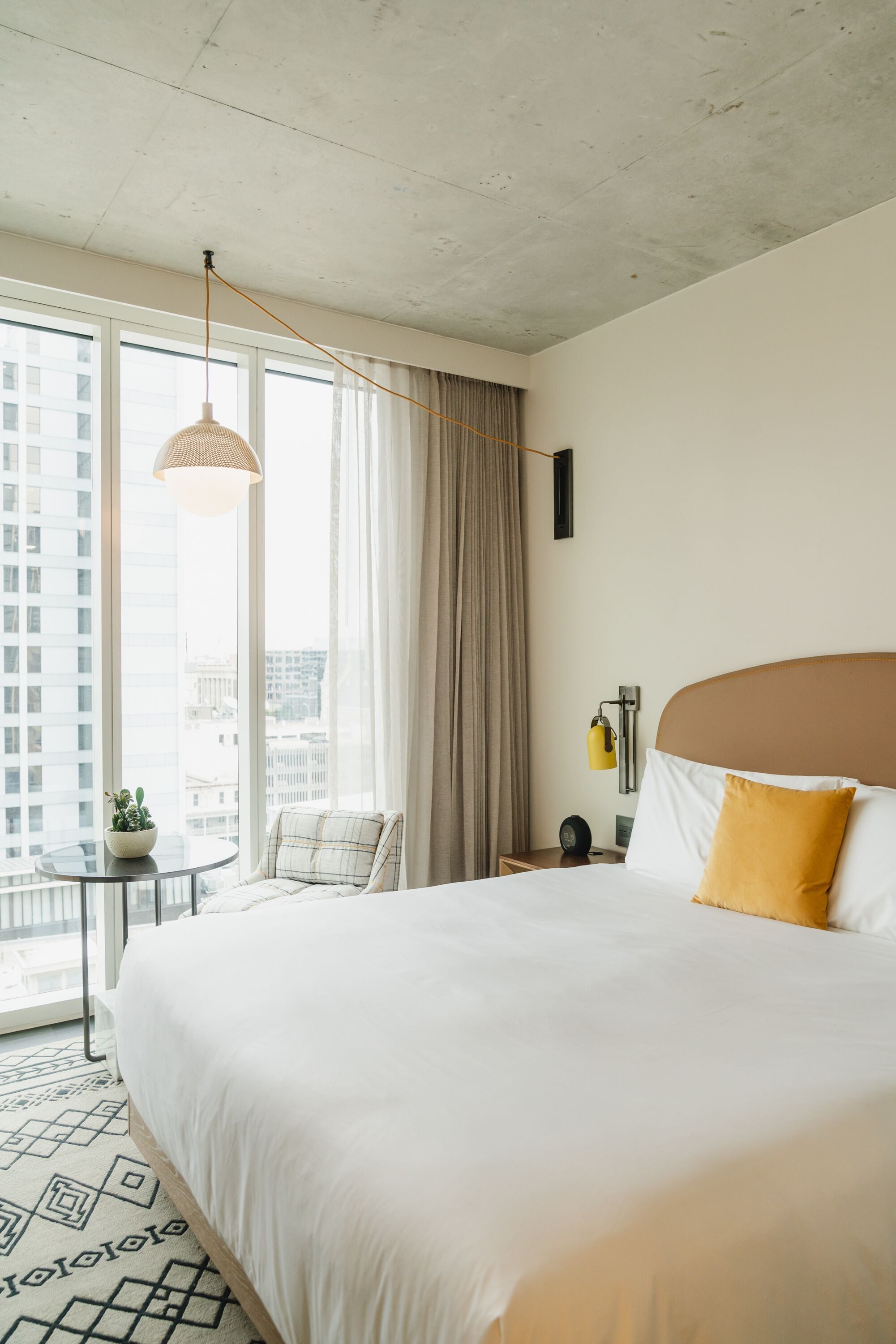 Quarto de hotel com uma cama grande, mesa de cabeceira com abajur, uma cadeira e vista da cidade através de grandes janelas.