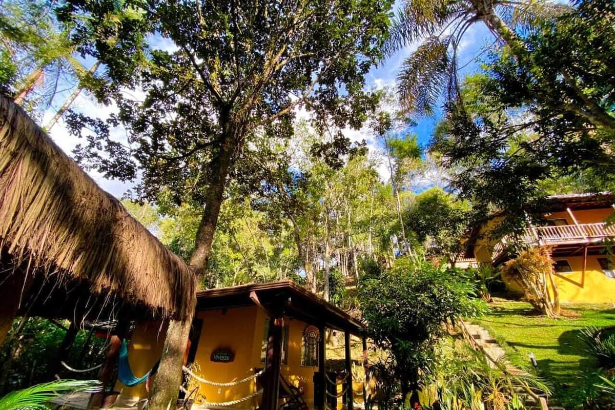 Uma cabana rústica com telhado de palha e uma pequena estrutura de madeira por perto, cercada por vegetação exuberante e árvores.