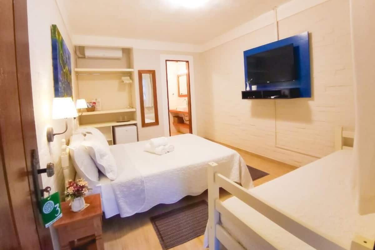 Quarto de hotel com duas camas, uma TV fixada na parede, uma mesinha com abajur e uma porta que dá para o banheiro.