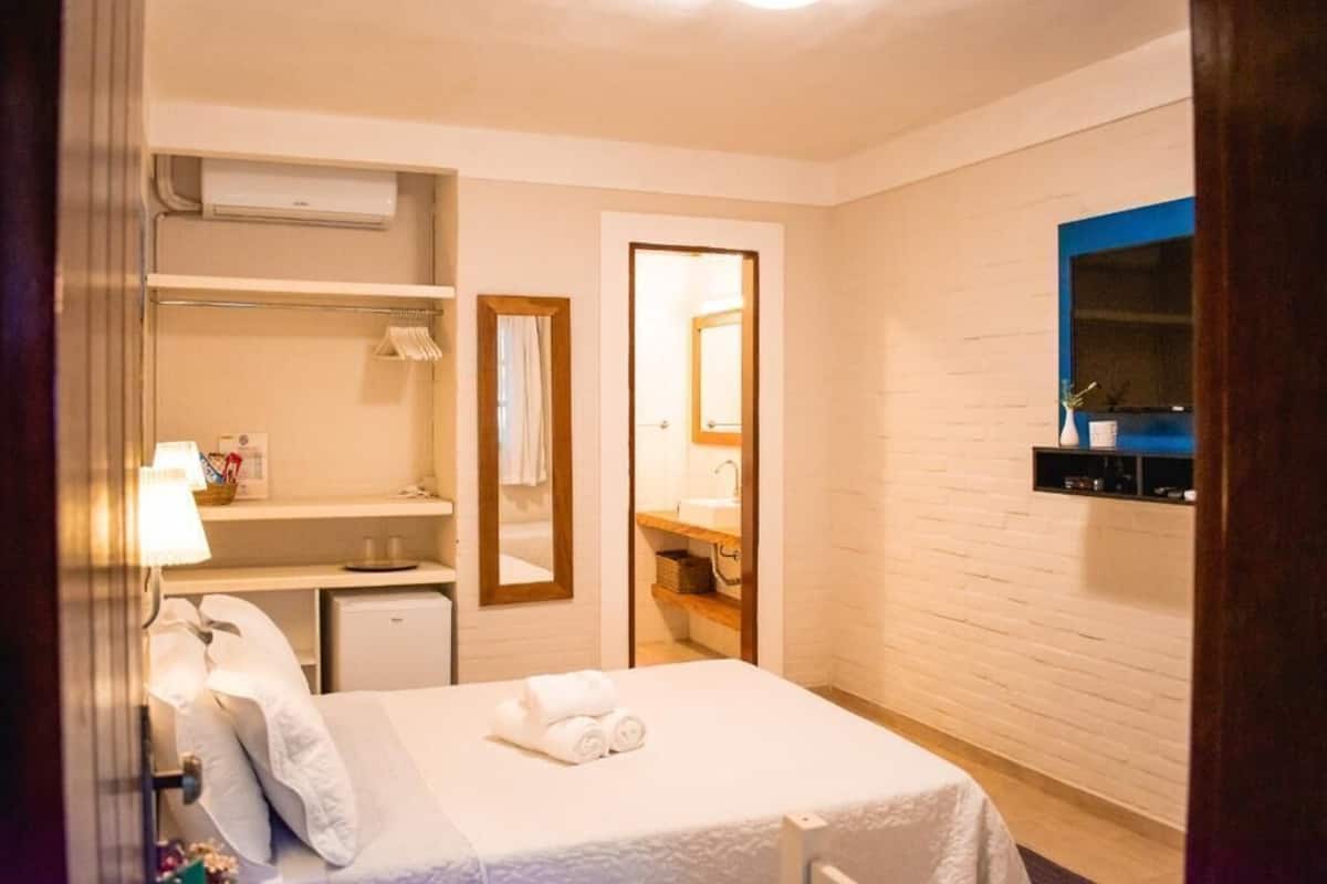 Quarto de hotel com cama, escrivaninha, espelho, prateleira com amenities de higiene pessoal e janela.