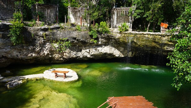 Uma piscina natural com um banco de madeira e uma pequena plataforma, rodeada por terreno rochoso e vegetação.