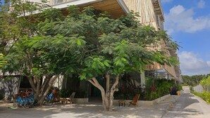 Um edifício com fachada moderna, uma área de estar externa coberta com cadeiras e uma mesa, e uma árvore ao primeiro plano.