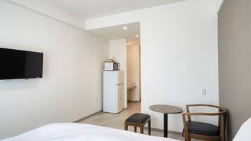 Quarto de hotel com cama, televisão fixada na parede, mesa pequena, duas cadeiras e frigorífico.