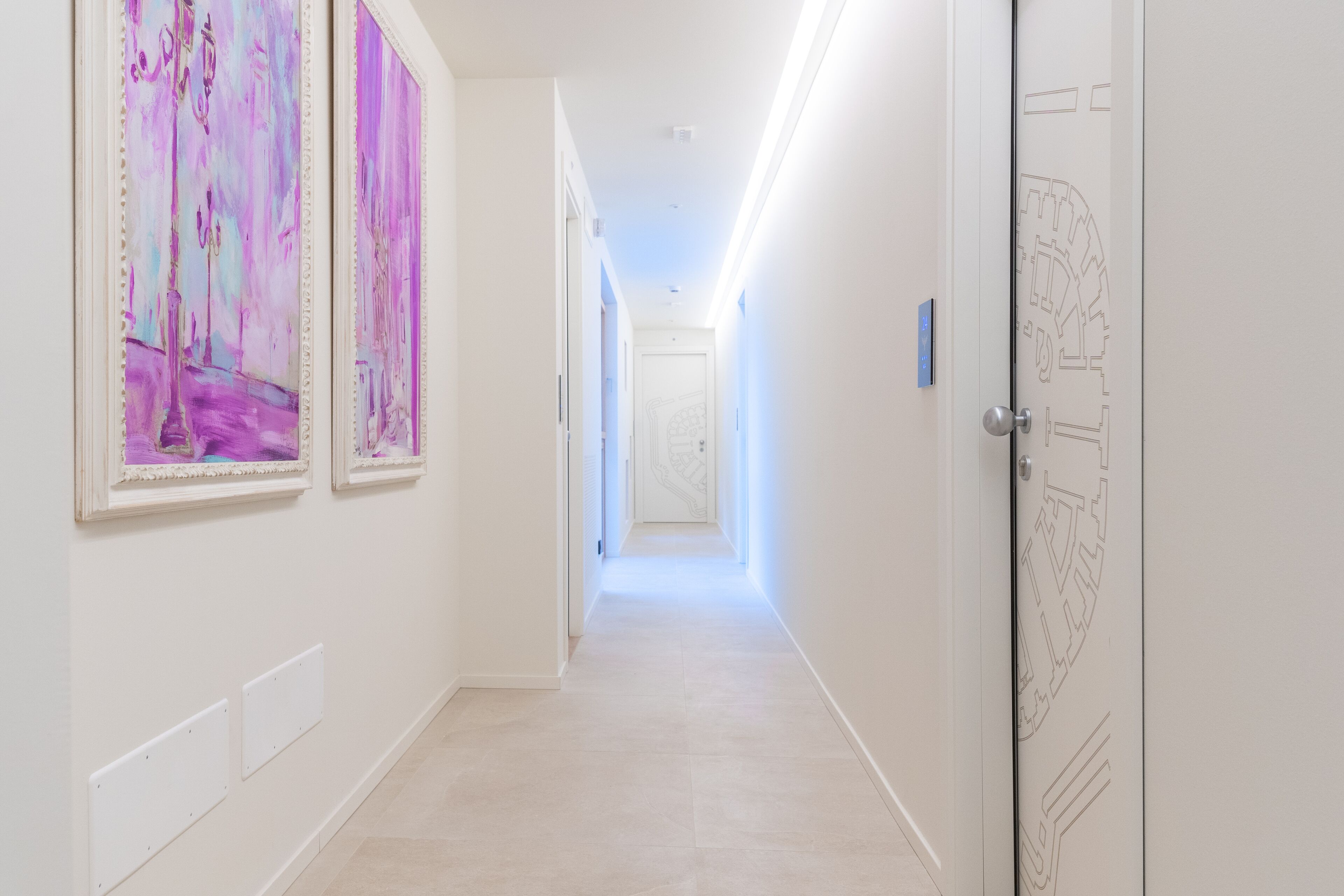 Um corredor com piso claro, paredes brancas e uma porta com design geométrico. Há duas obras de arte emolduradas na parede esquerda.