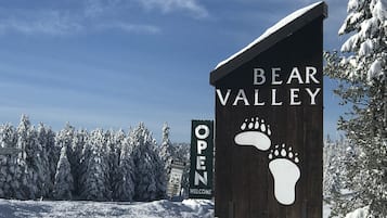 Uma placa de madeira com a inscrição 'BEAR VALLEY OPEN' e um símbolo de pata, rodeada por árvores cobertas de neve.