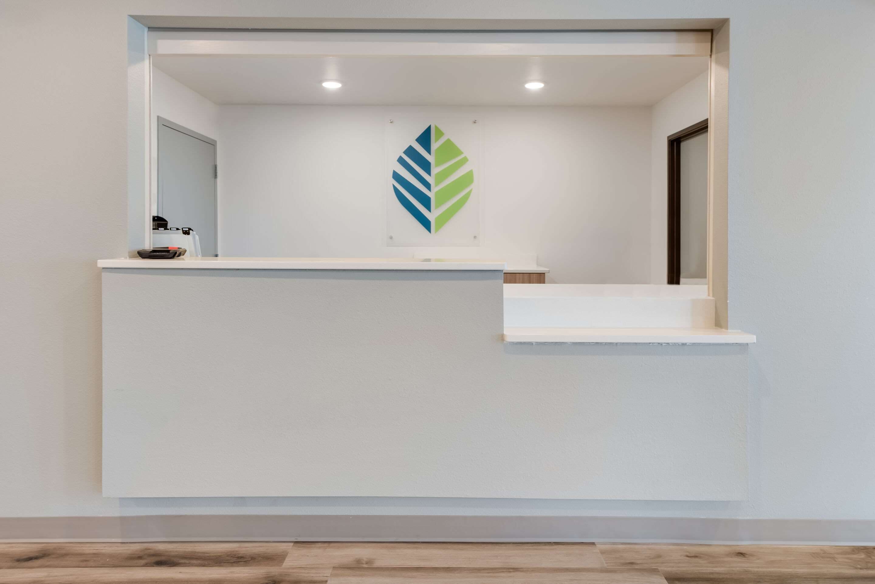 Uma área de recepção com um balcão branco, um logotipo verde e azul na parede e uma porta à esquerda.