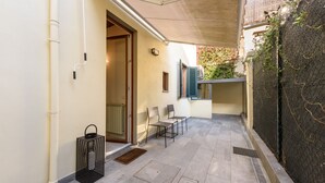 Uma área externa coberta com chão de azulejos, uma porta que dá para o interior e um pequeno espaço de estar com cadeiras.