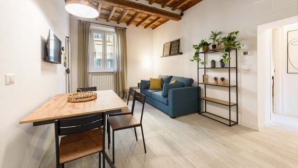 Uma sala de estar moderna com uma mesa de jantar de madeira, sofá azul e uma estante de metal preto com plantas.