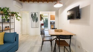 Uma sala de estar moderna com sofá azul, mesa de jantar de madeira e cadeiras pretas. Há uma TV fixada na parede e um ventilador de teto.