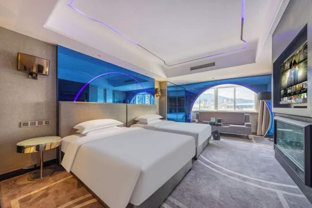 Quarto de hotel com duas camas, televisão, frigobar e uma janela grande.