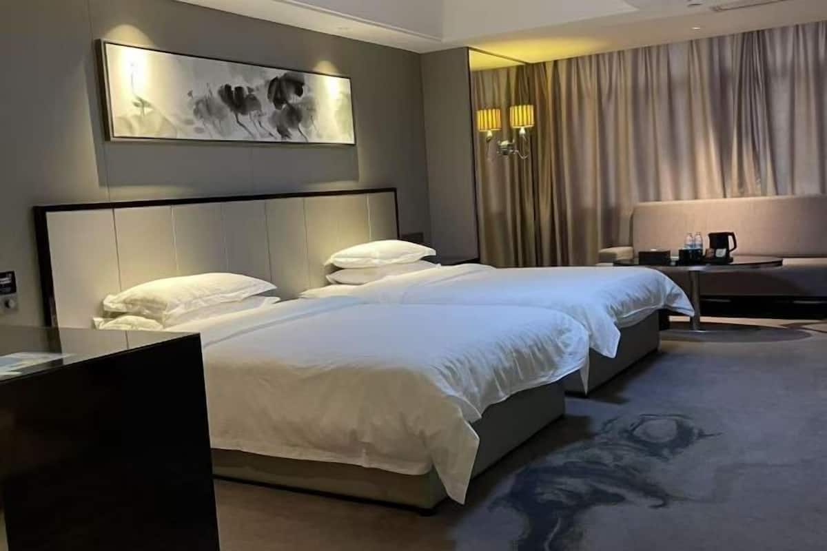 Quarto de hotel com duas camas, um quadro abstrato grande, uma mesa de cabeceira com abajur e um sofá.