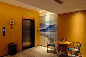 Um hall de hotel com parede de madeira, um elevador, uma pintura de montanhas e duas cadeiras com uma mesinha.