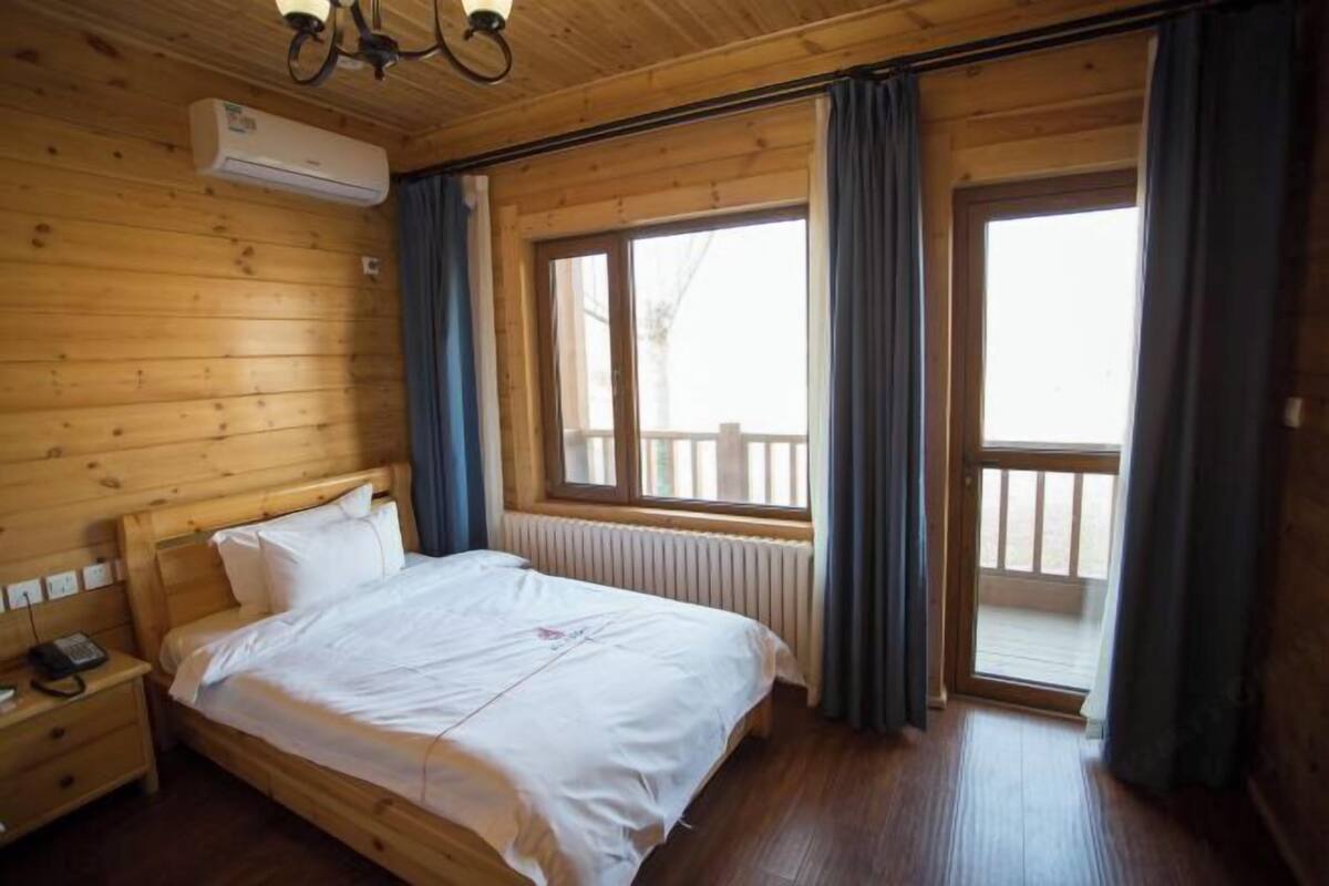 Quarto de cabana de madeira com cama, mesa de cabeceira, aparelho de ar condicionado e janela com cortinas.
