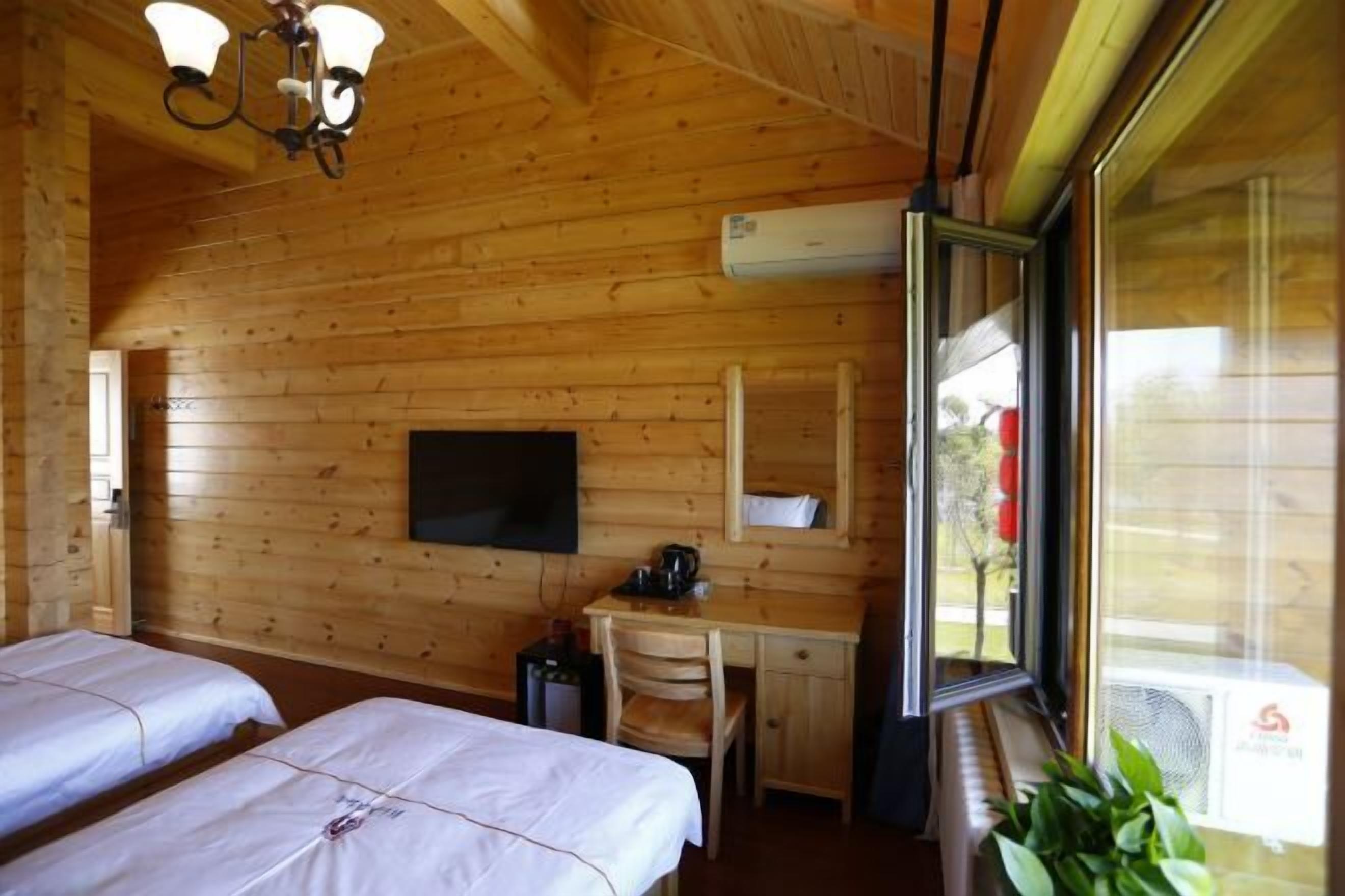 Quarto de cabana de madeira com duas camas, uma escrivaninha, uma cadeira, televisão e janela.