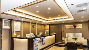 Uma área de recepção de hotel com design moderno, com balcão de recepção, espaço de estar com sofá e mesa de centro, e um relógio fixado na parede.