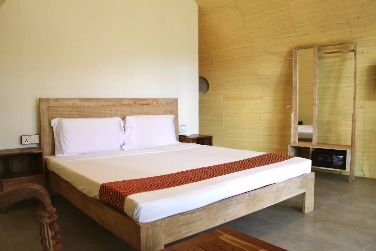 Uma cama de madeira com roupa de cama branca e um colcha vermelha estampada num quarto com paredes e chão de madeira.