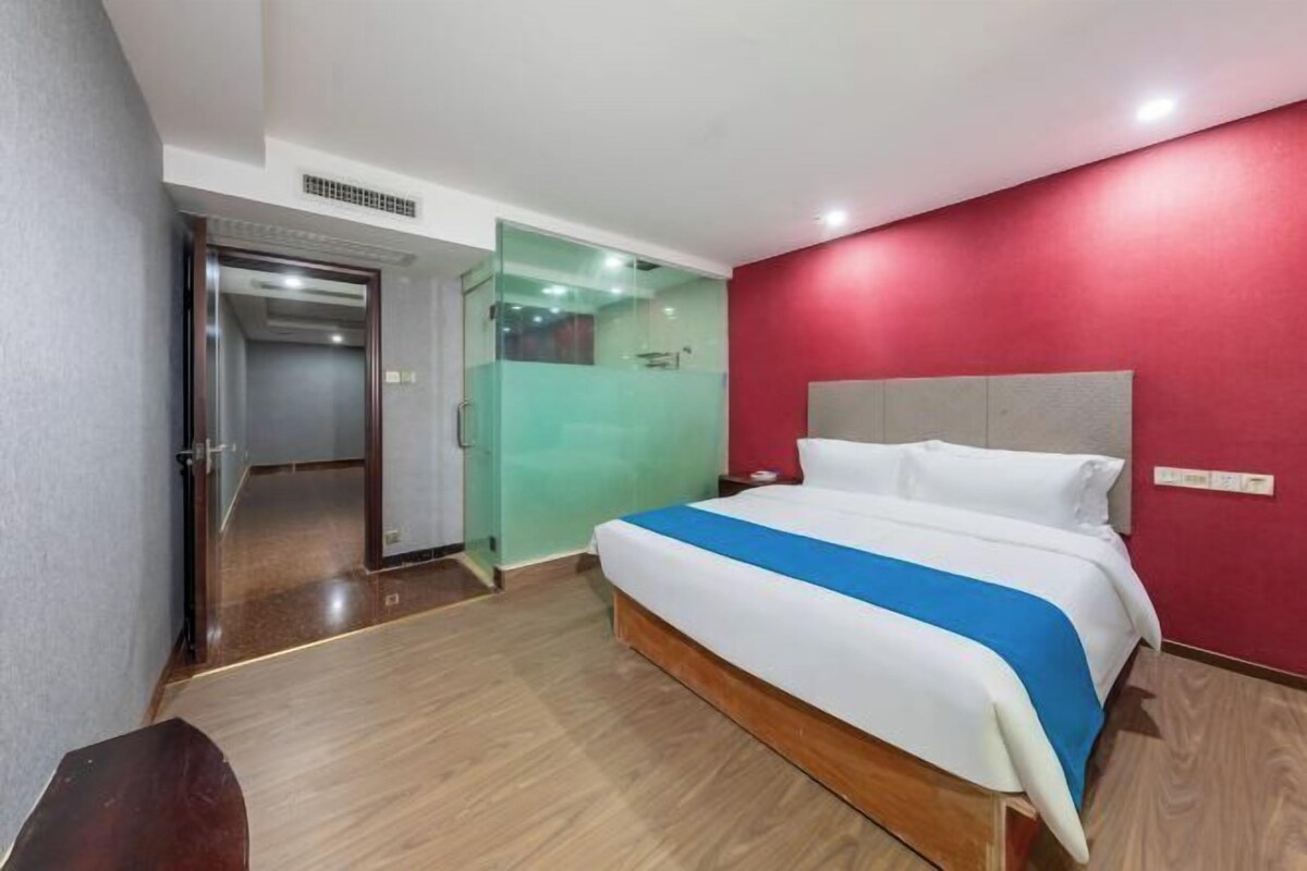 Quarto de hotel com cama, parede de destaque vermelha, piso de madeira e box de vidro.