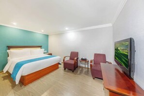 Quarto de hotel com uma cama, duas poltronas, uma mesinha e uma televisão na parede.