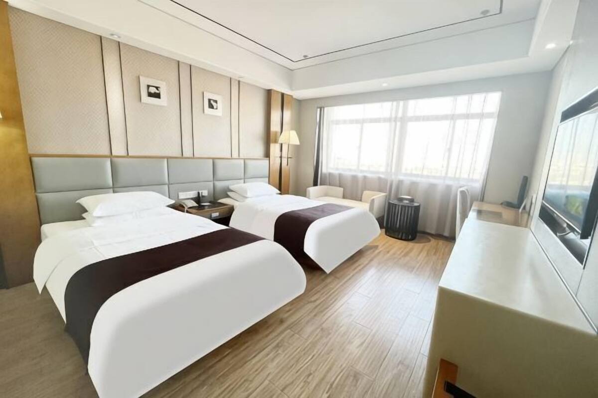 Quarto de hotel com duas camas, uma escrivaninha, uma cadeira, uma televisão e uma janela grande.