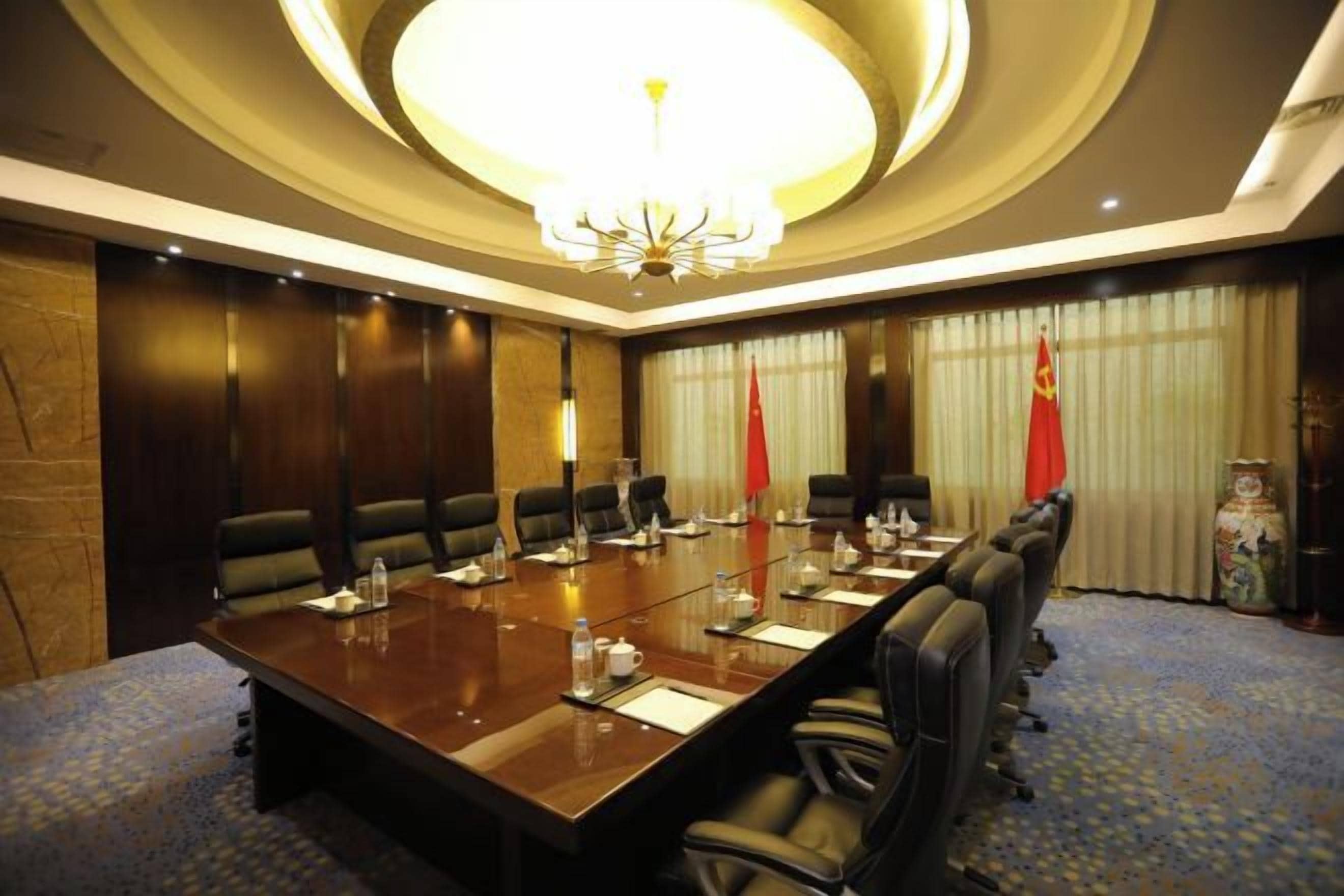 Uma sala de conferência com uma mesa comprida, cadeiras, um candelabro, bandeiras e um vaso.